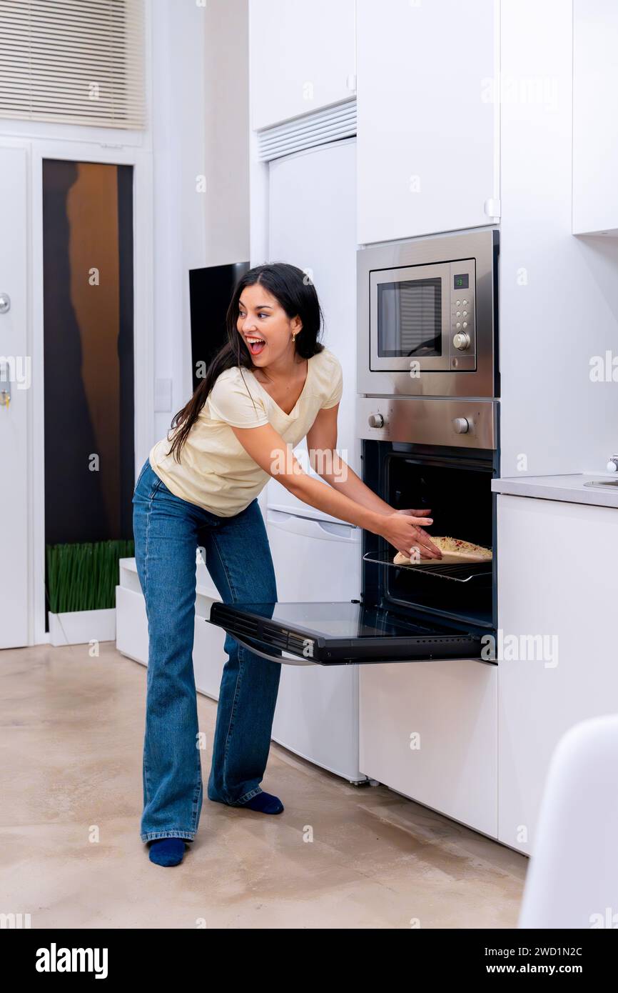 Hispanic woman happily slides a pizza into the oven in a modern white ...