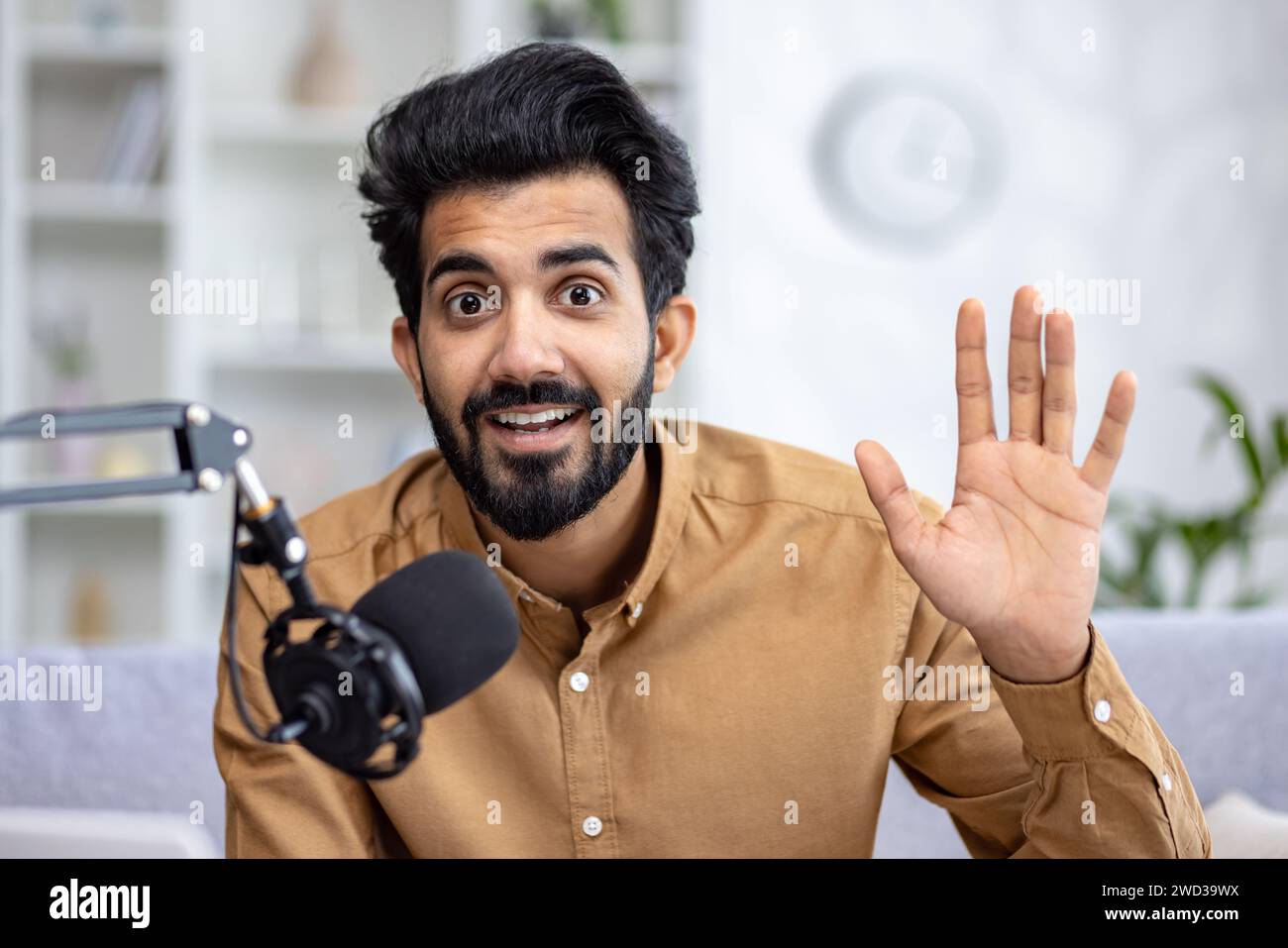 A cheerful young Indian man waving while talking into a microphone ...