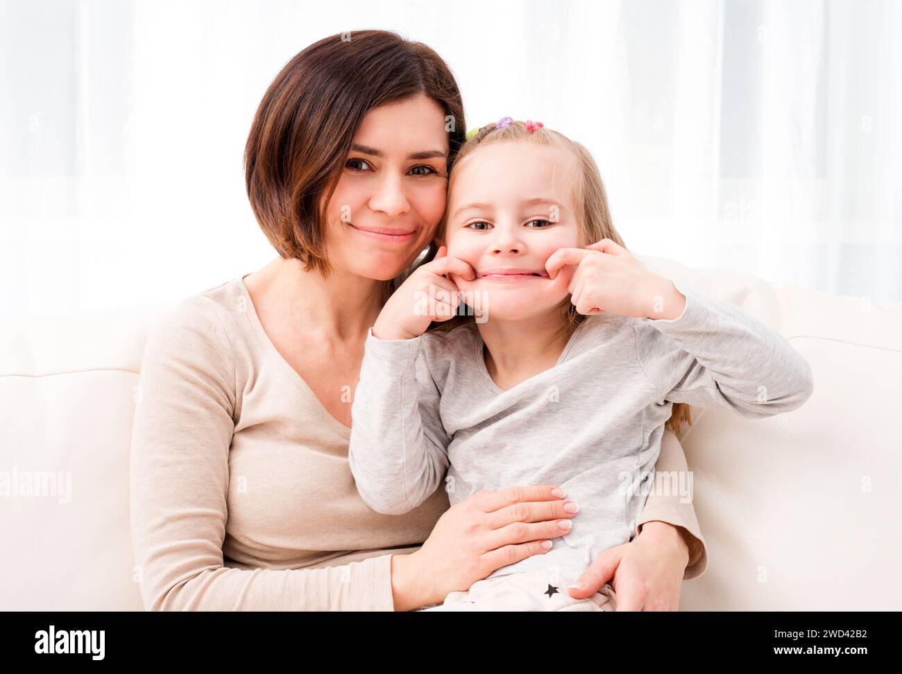 Cheerful little girl sitting with mum on the coach and making funny ...