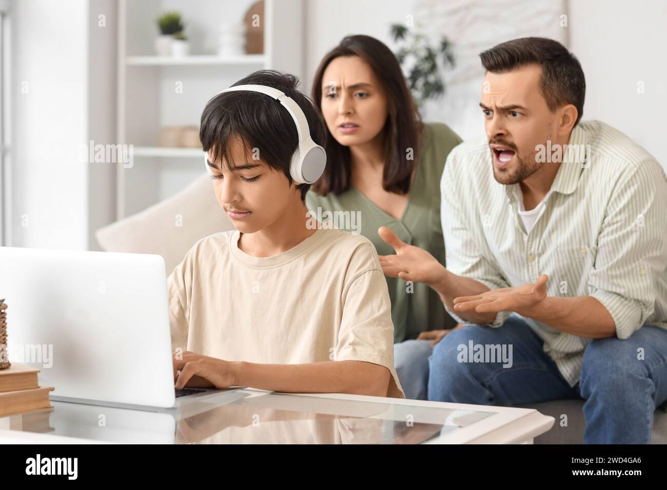 Teenage boy in headphones using laptop and his angry parents at home ...