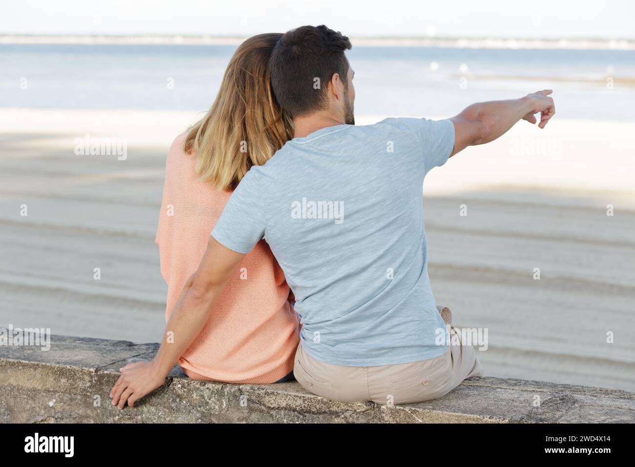 couple sat on wall overlooking beach and pointing into distance Stock ...
