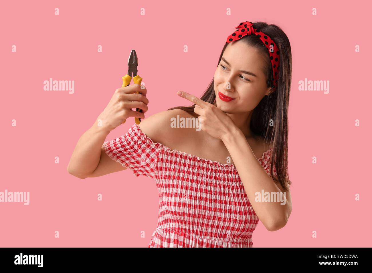 Portrait of young pin-up woman pointing at pliers on pink background ...