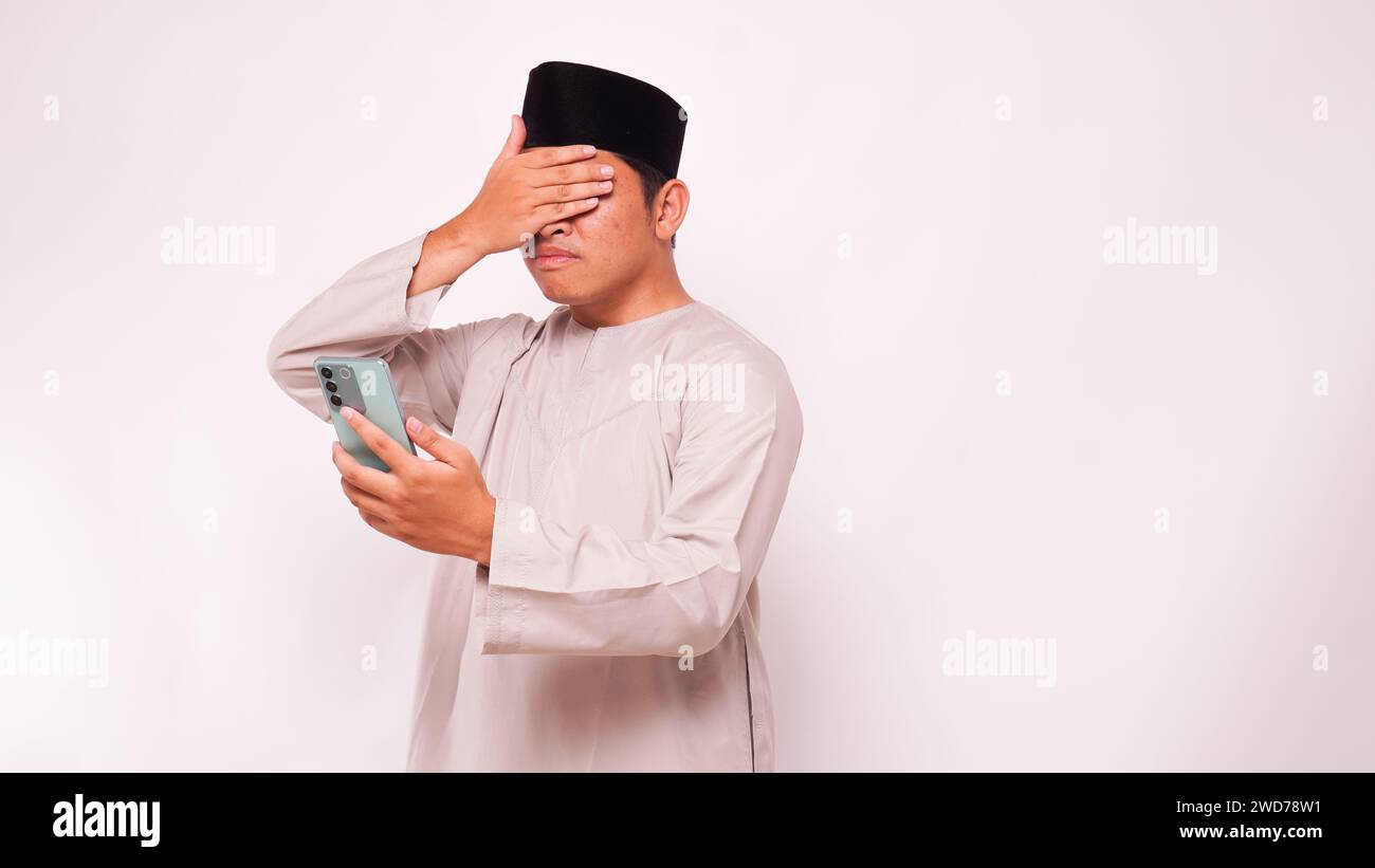 Asian muslim man covering his eyes while looking at smartphone during ...