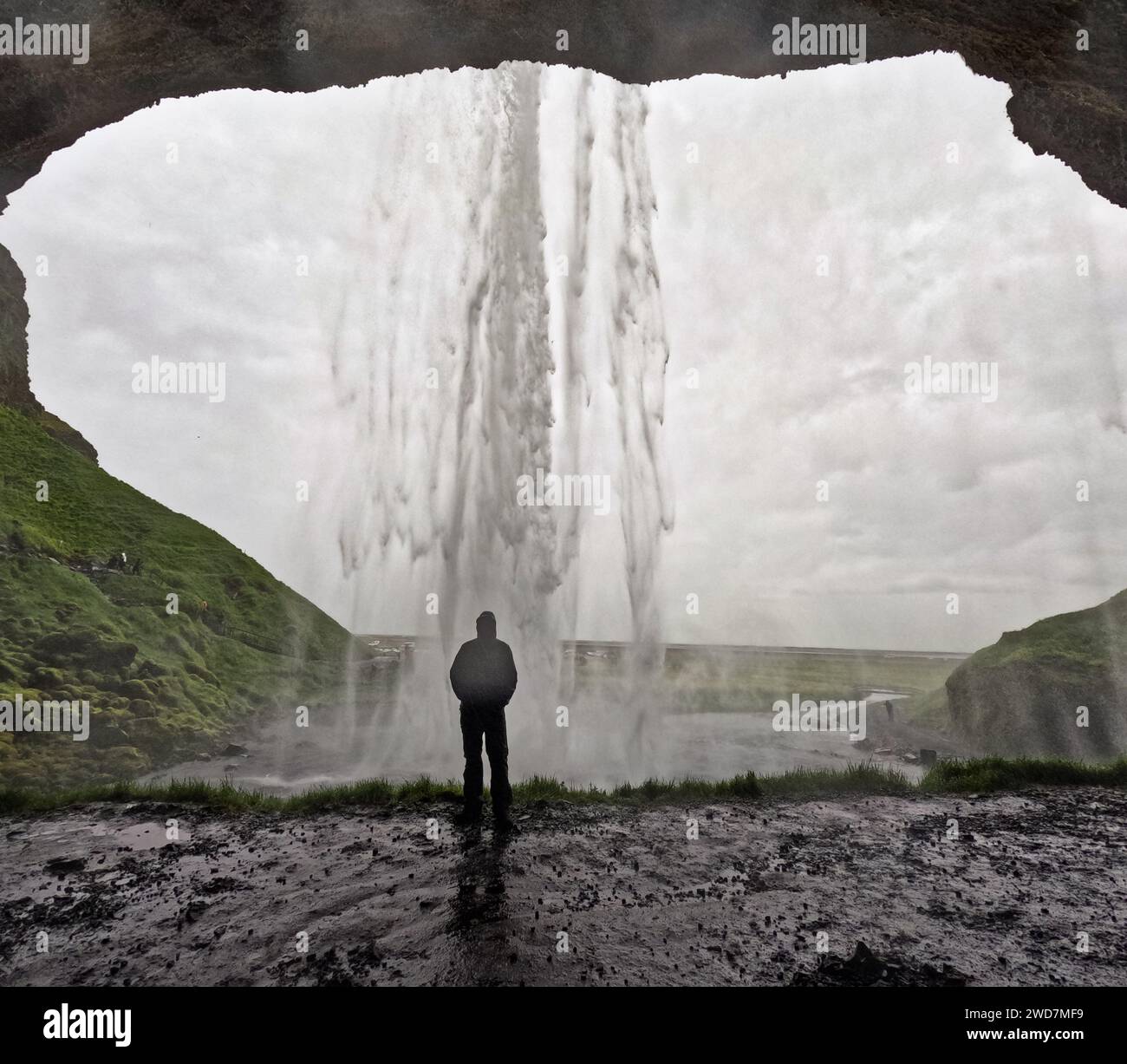 Man stands under waterfall in mouth of cave with cloudy sky Stock Photo ...