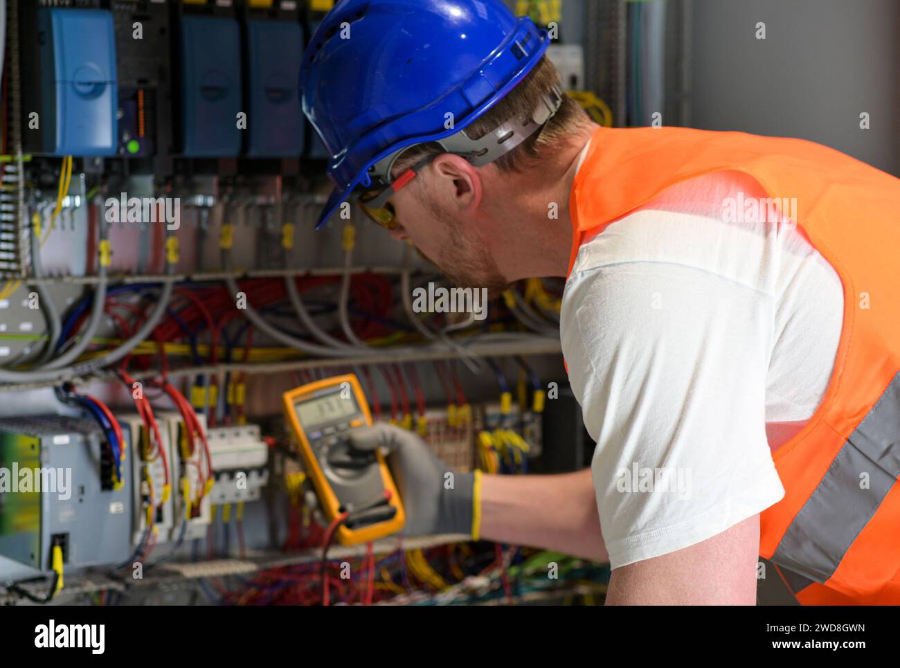electrician in blue helmet, yellow glasses and orange vest measures ...