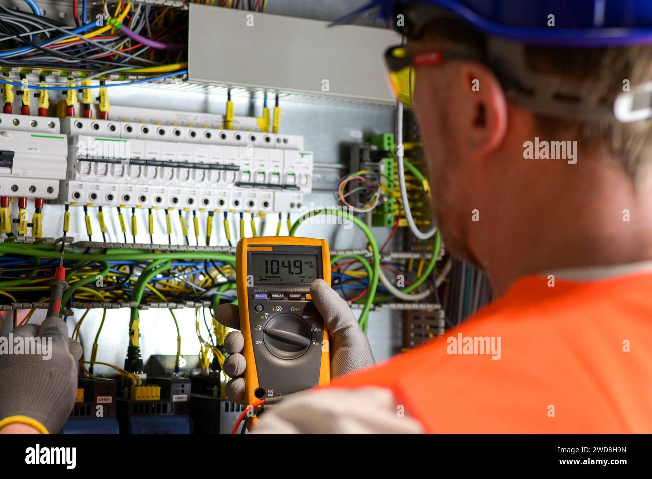electrician in blue helmet, yellow glasses and orange vest measures ...