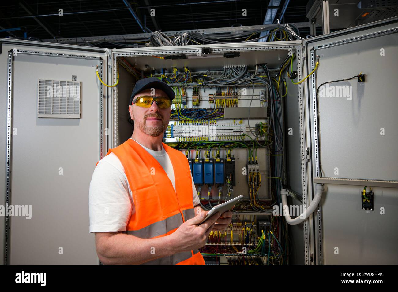 an electrician in a cap and orange vest installs a large electrical substation using a tablet ...