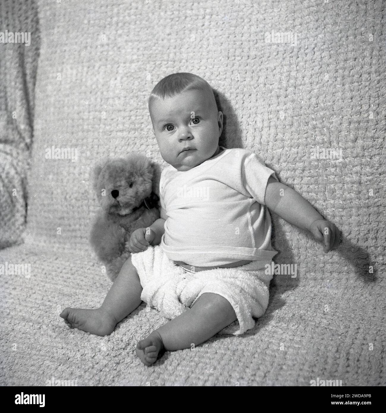 1960s, historical, a baby boy with his teddy bear beside him sitting on ...