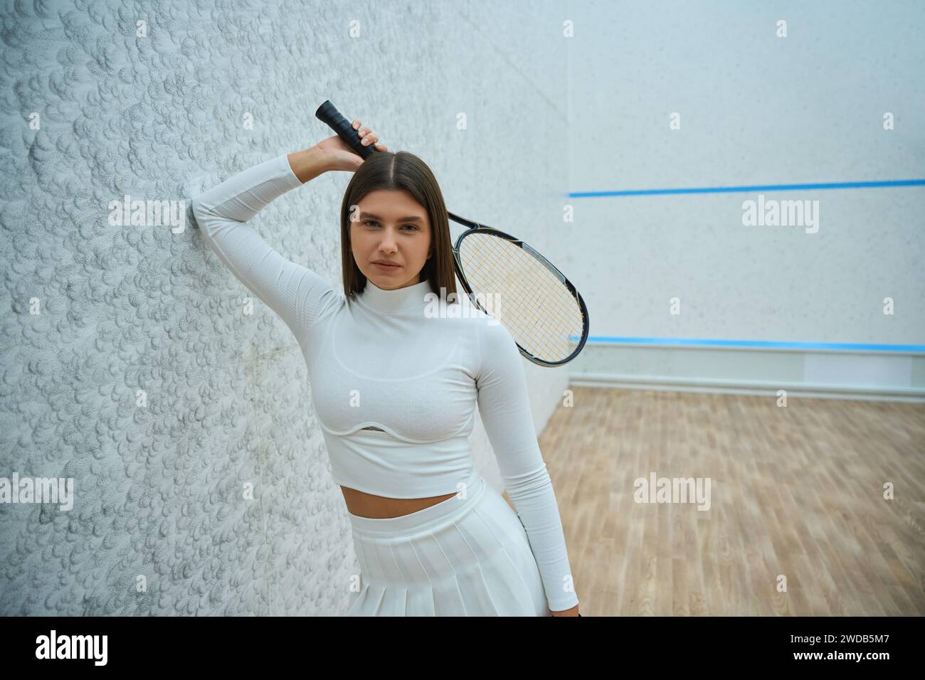 Sporty beautiful woman in white attire holding squash racket leaning on ...