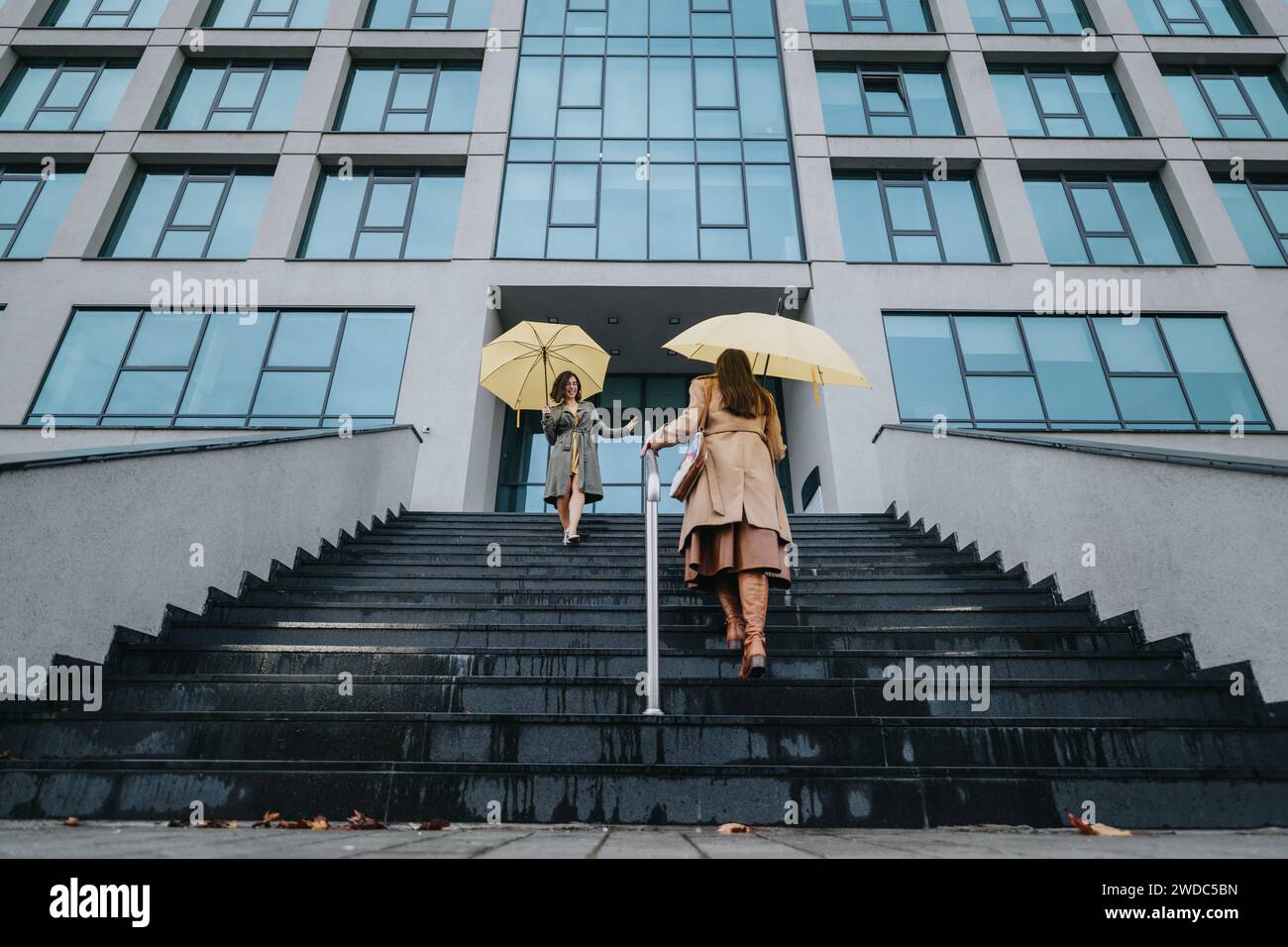 Two women holding yellow umbrellas walking down office building steps ...