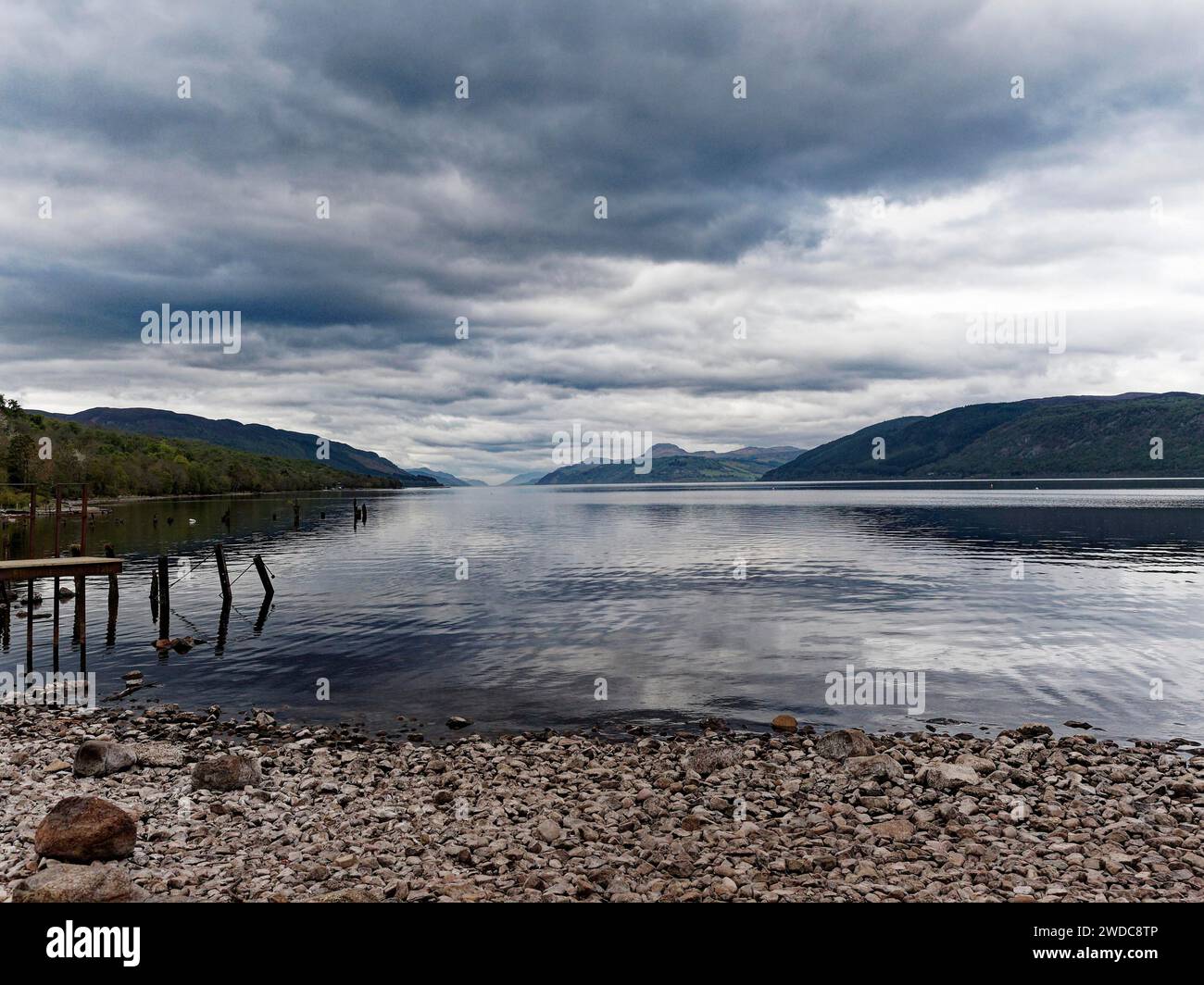 A tranquil loch with a pebble beach and pier under a cloudy sky, on the ...