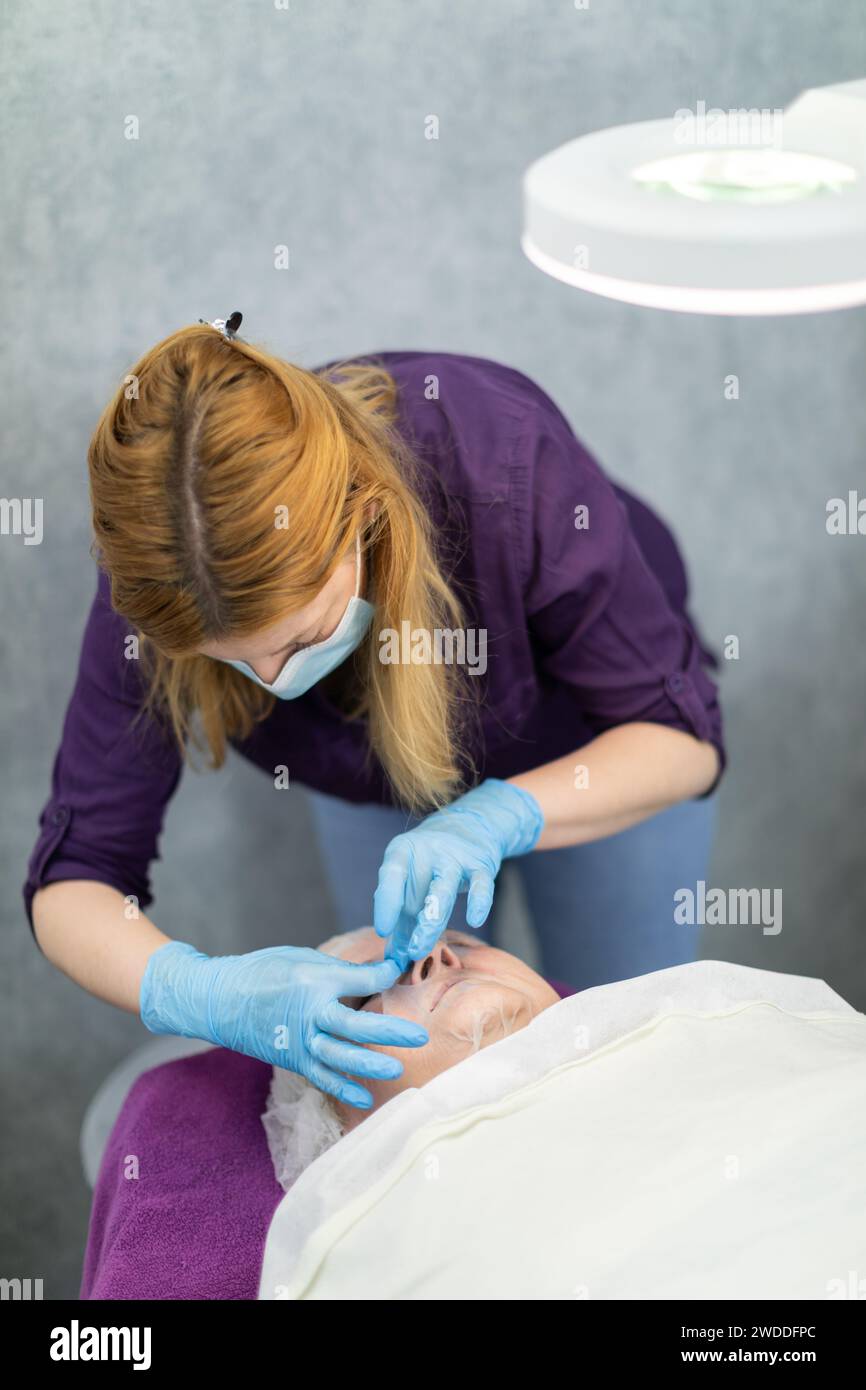 The cosmetologist is wearing a purple smock, surgical mask and ...