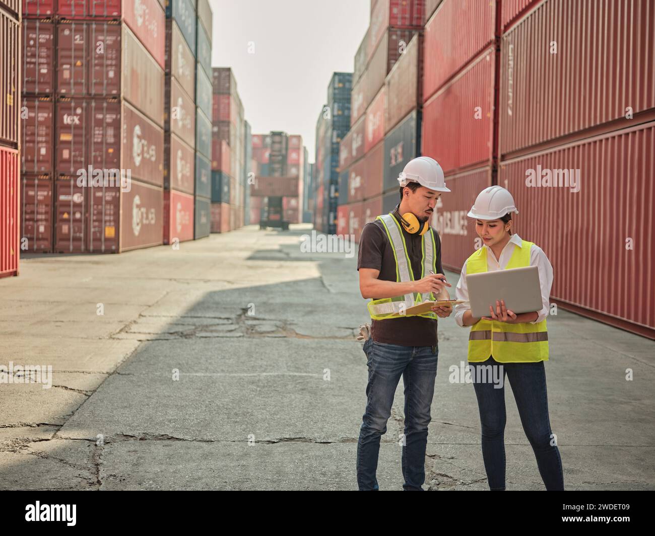 young Asian woman and man engineer working with co-worker at overseas shipping container yard ...