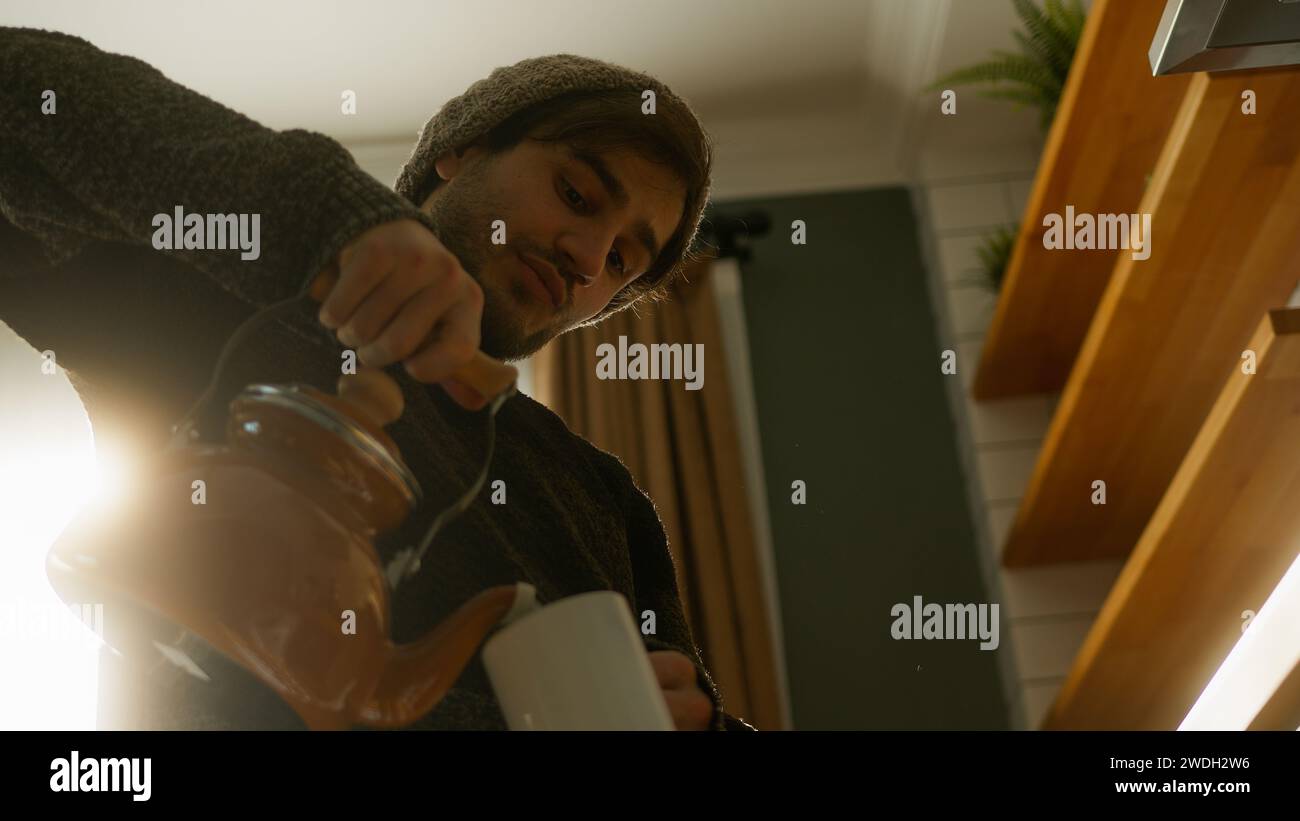 Young man with beanie stands in the modern kitchen, pours tea from a ...