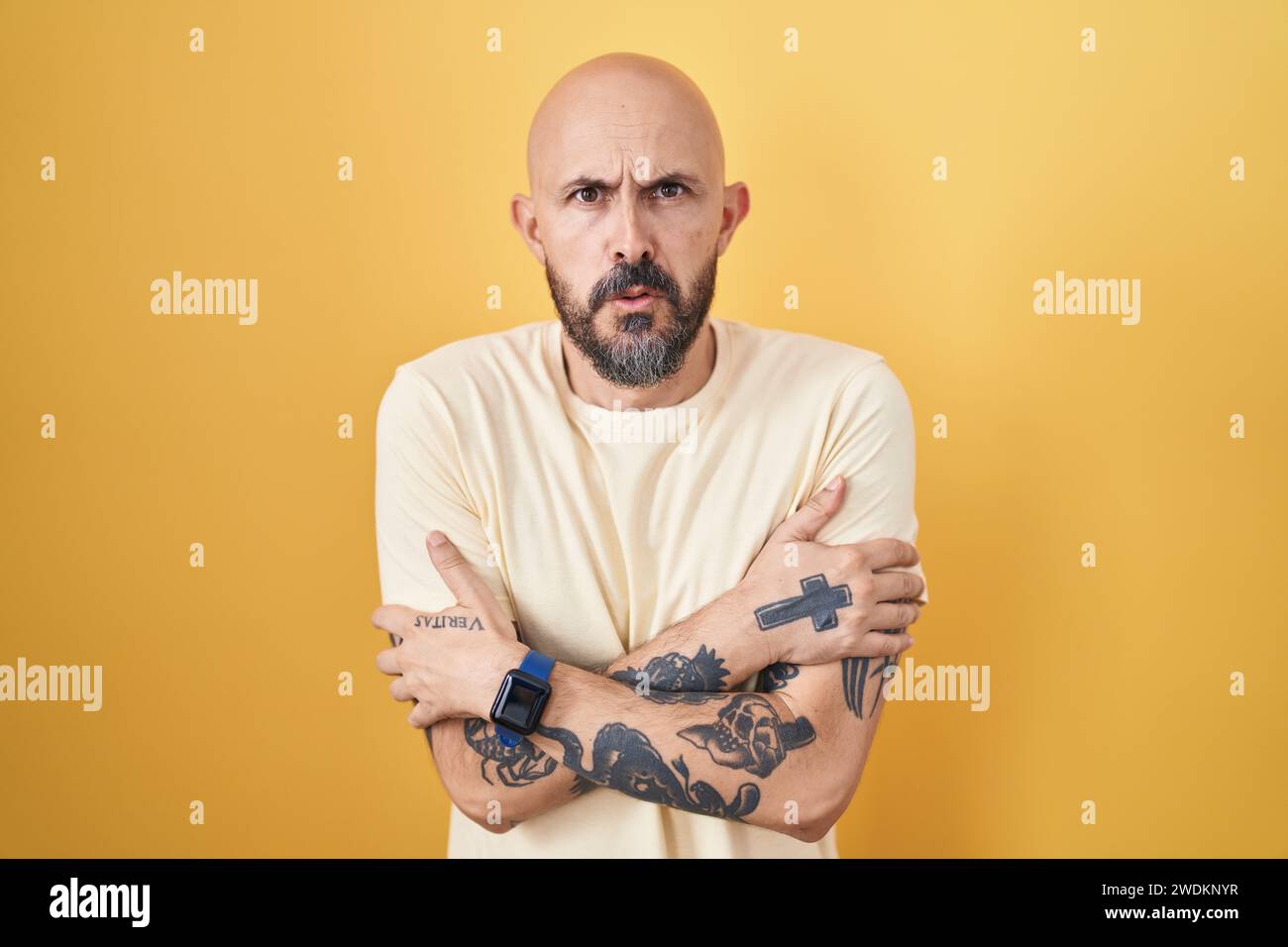 Hispanic man with tattoos standing over yellow background shaking and ...