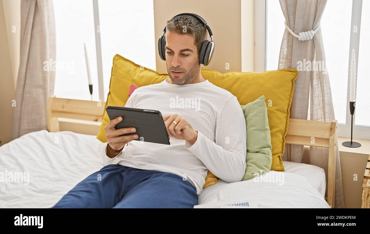 Relaxed man with headphones using tablet on bed in a bright, cozy ...