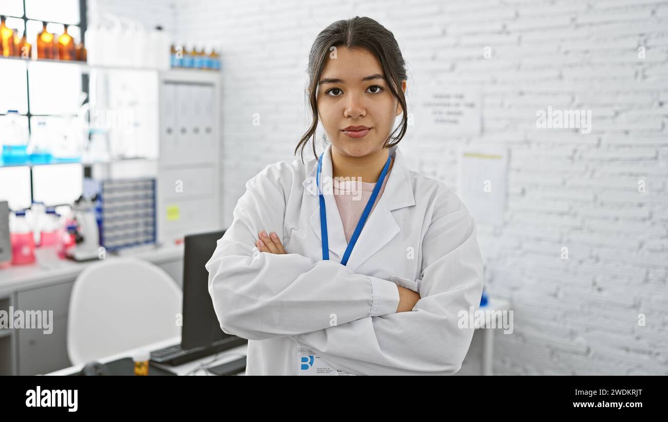 Confident young hispanic woman in lab coat with arms crossed standing ...