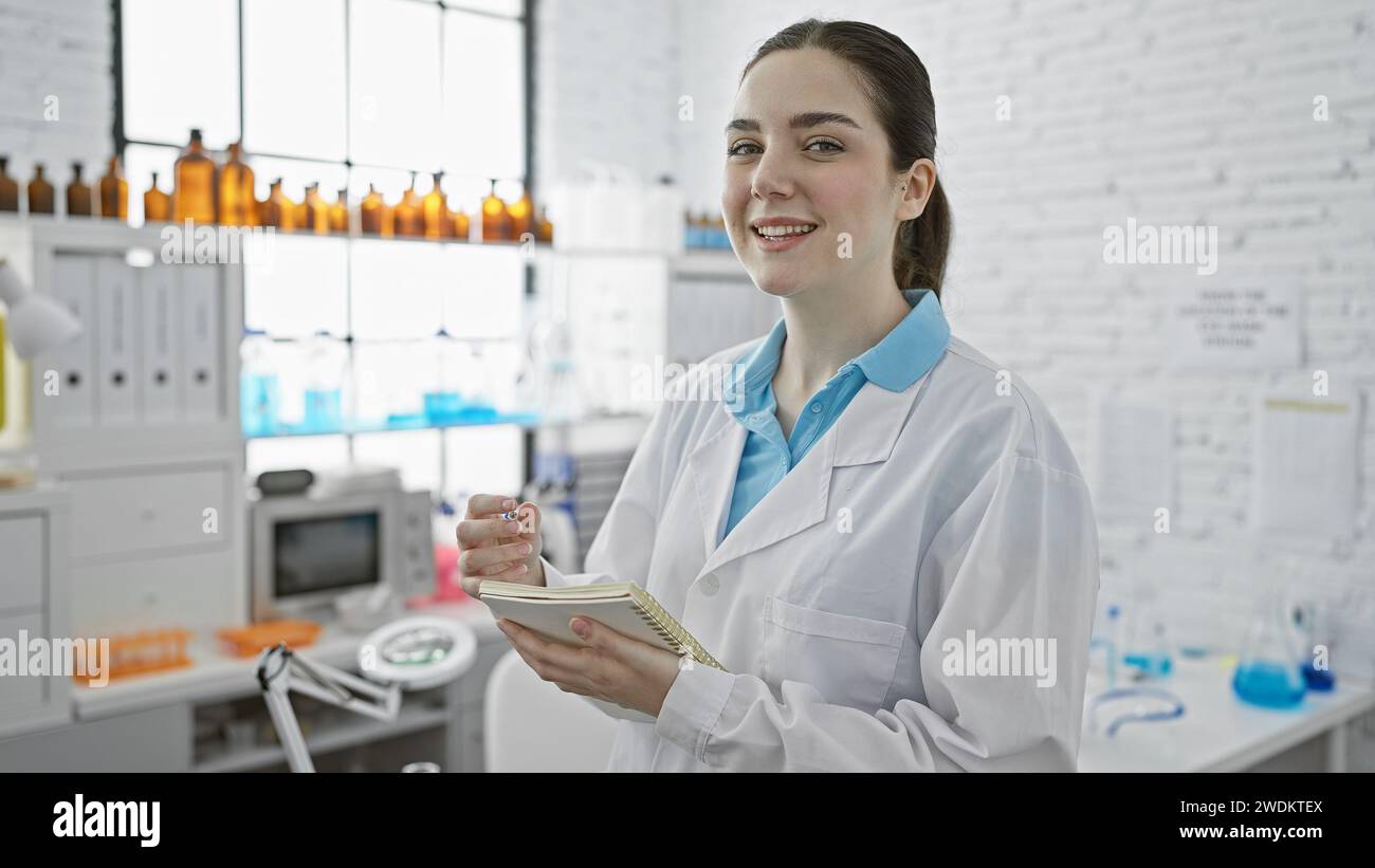 Confident young woman in a lab coat holding a notebook inside a modern ...