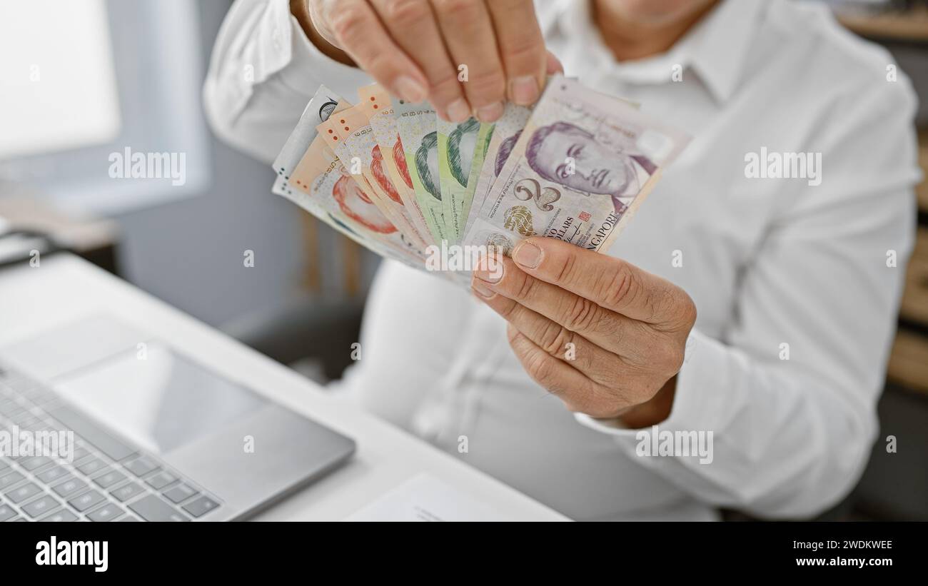 A middle-aged man in an office holds singaporean currency, portraying ...