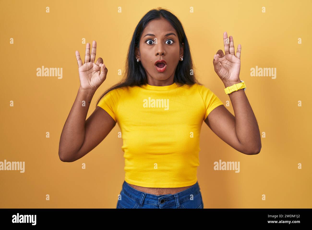 Young indian woman standing over yellow background looking surprised ...