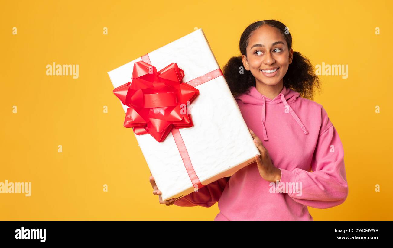 Curious black teenager girl shaking large box checking gift, studio ...