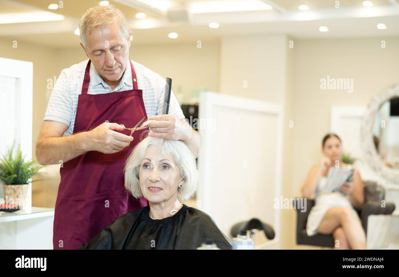 Experienced senior man master makes performs haircut for mature woman ...