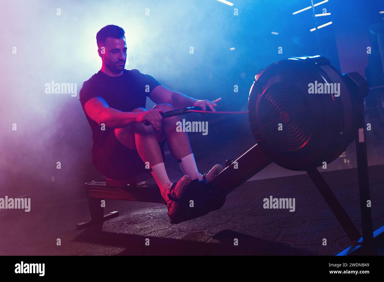 A male athlete pulls the oars on a rowing machine, his form lit by ...