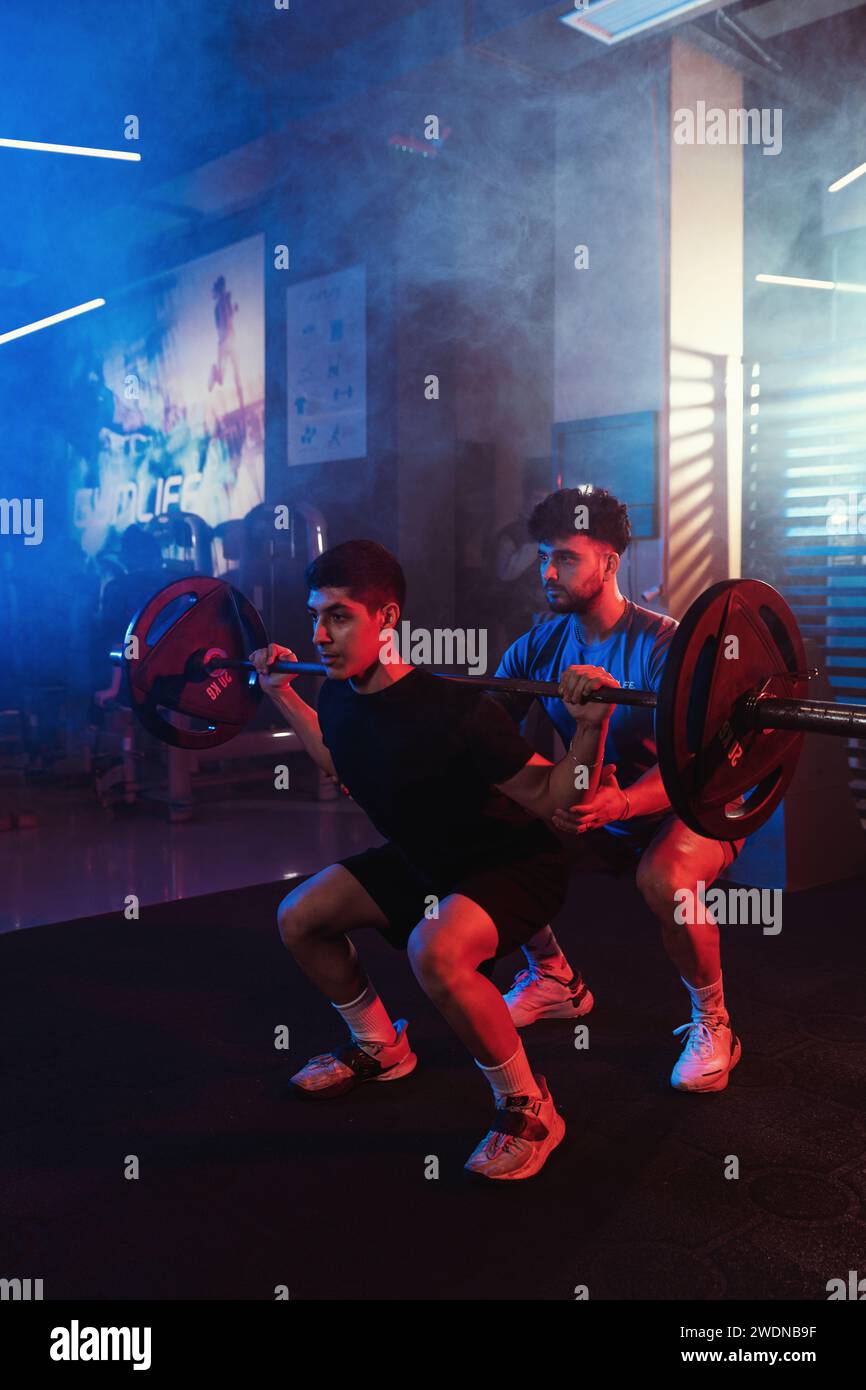 A focused male athlete performs squats under the guidance of a trainer ...