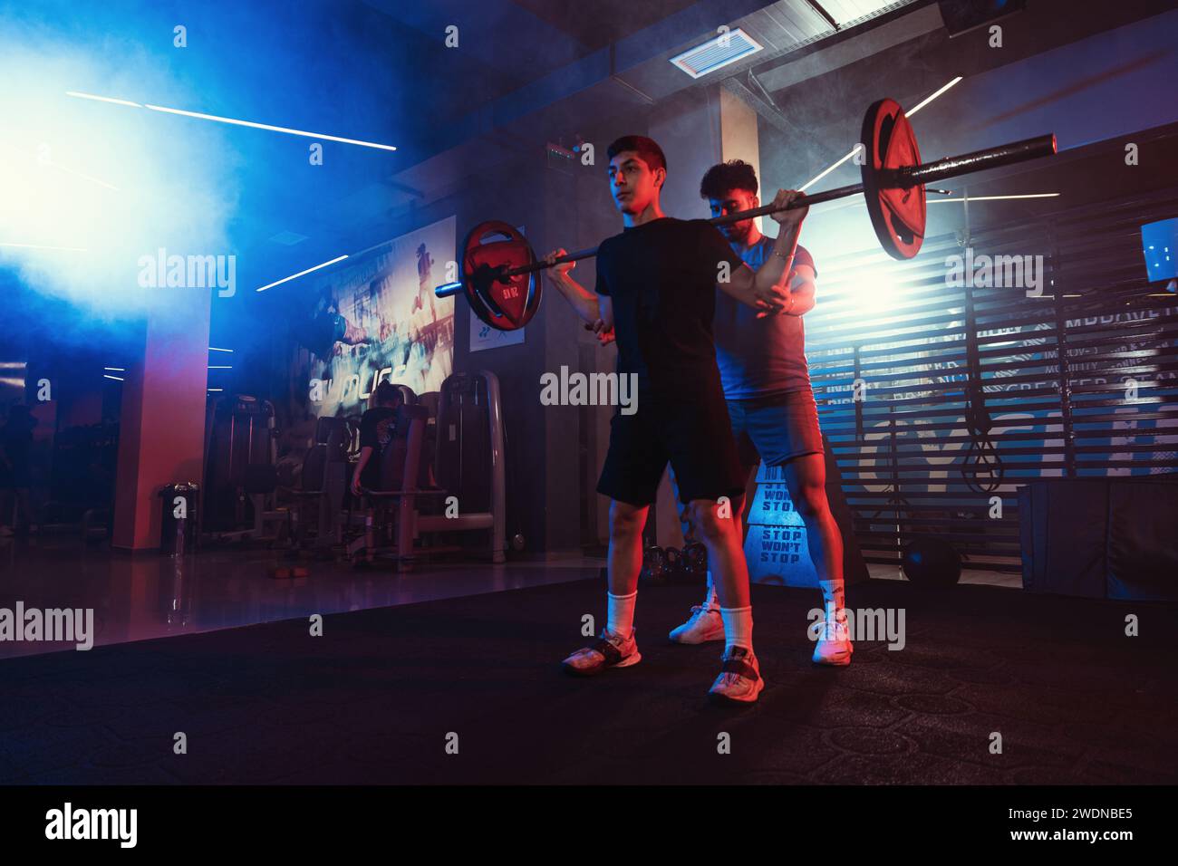 A focused male athlete performs squats under the guidance of a trainer ...