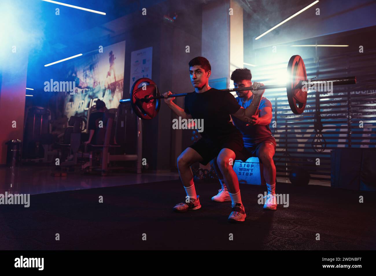 A focused male athlete performs squats under the guidance of a trainer ...