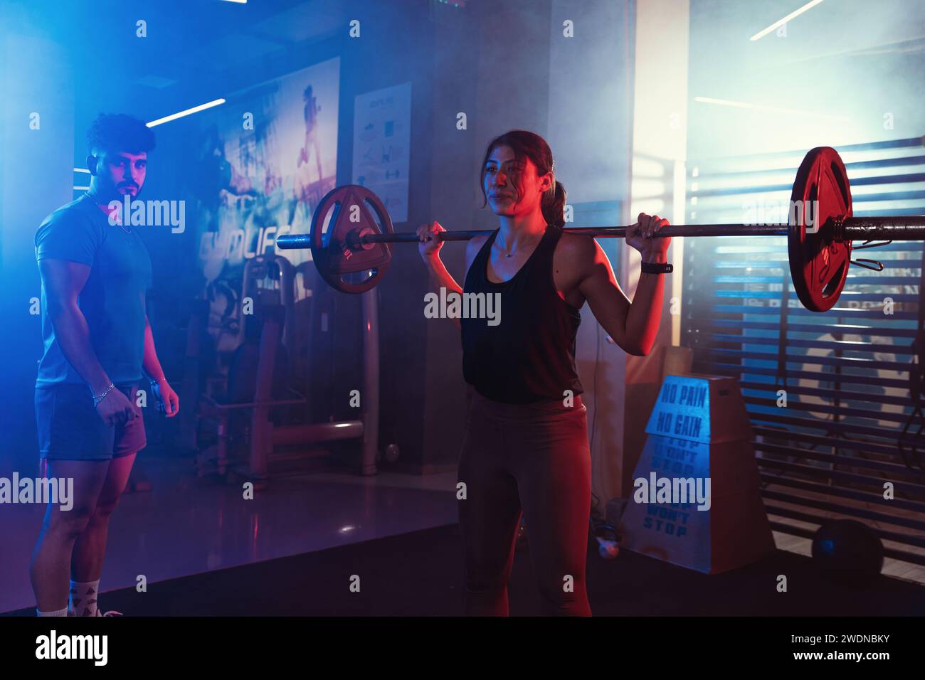 Under the watchful eye of her trainer, a female athlete squats with a ...