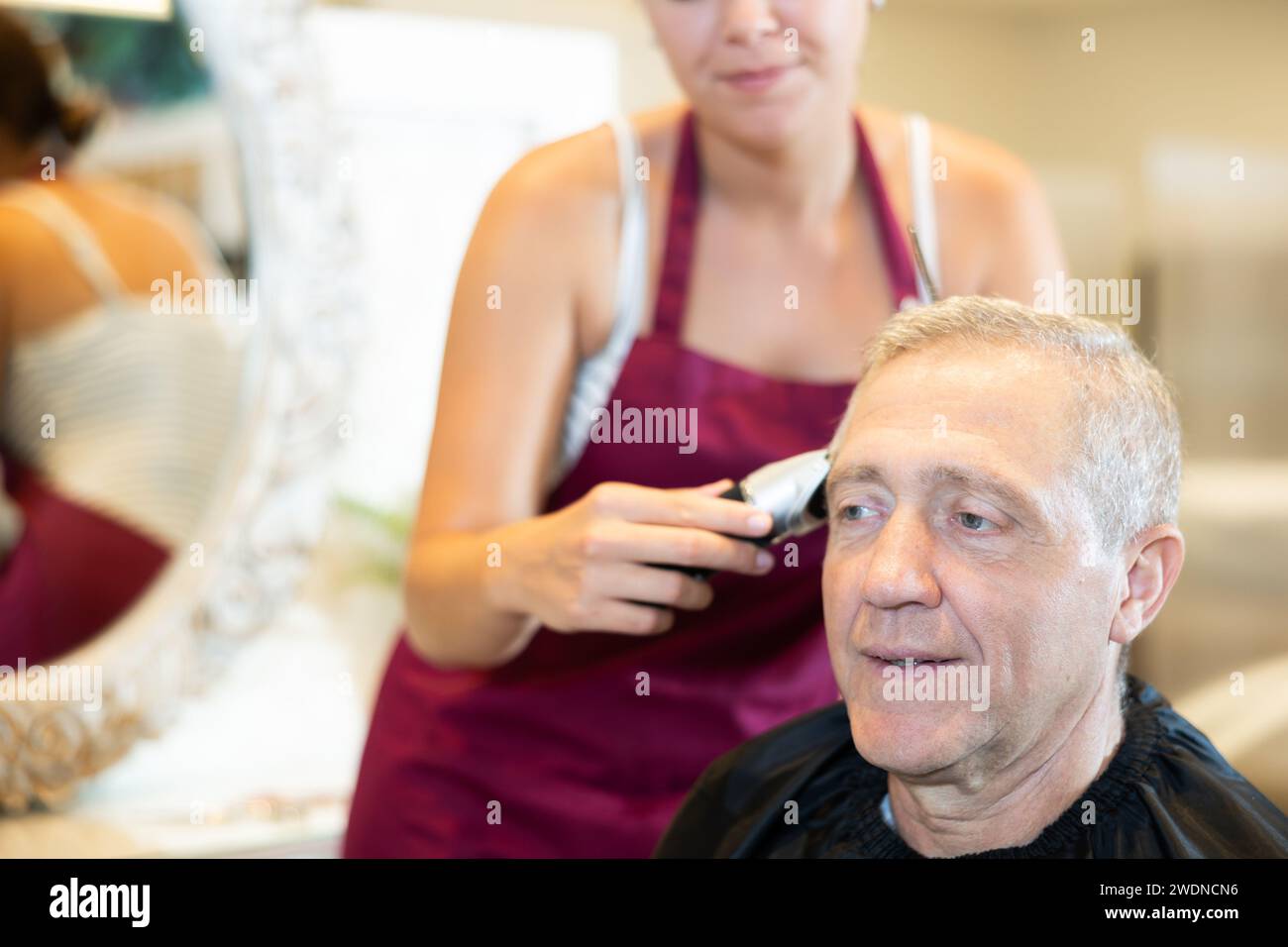 Cropped young woman hairdresser performs haircut for elderly man using ...