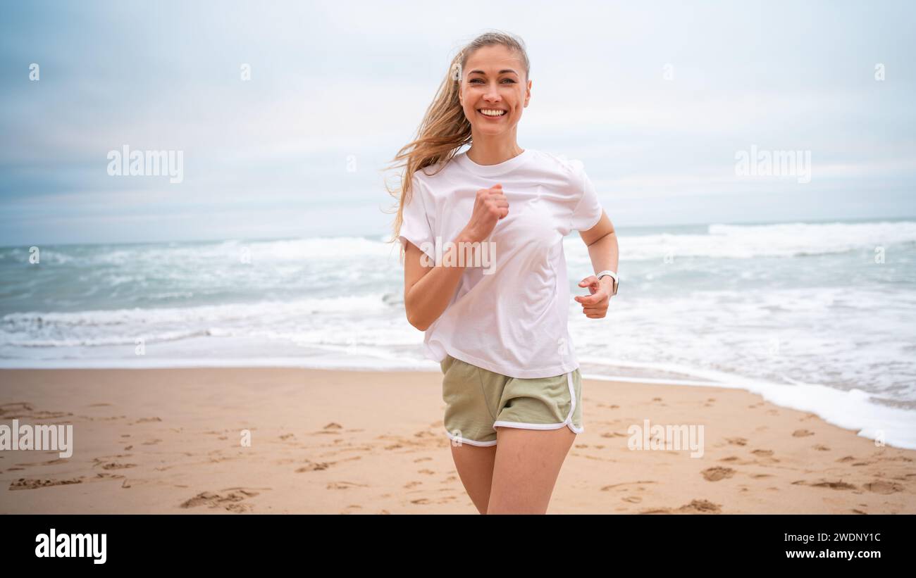 Smiling female athlete jogging on sandy sea beach. Fit woman in white ...