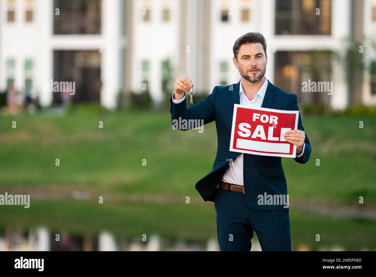 Successful real estate agent in a suit holding for sale sign near new ...