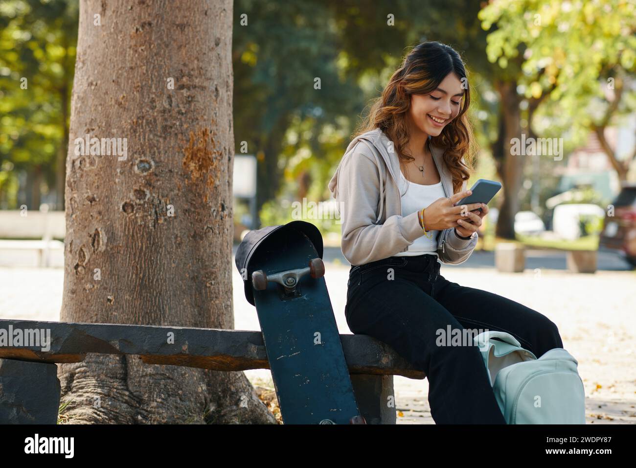 Joyful girl sitting on bench next to skateboard and texting friends ...