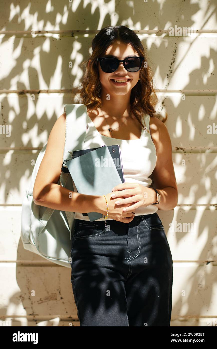 Cheerful college student with textbooks standing at wall with tree ...