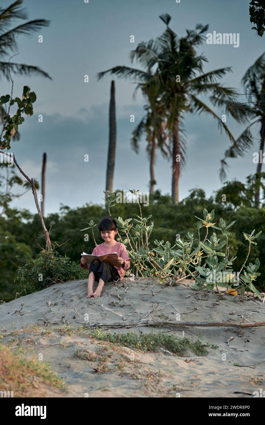 A cute little girl sits under a tree reading a book in the tropical ...
