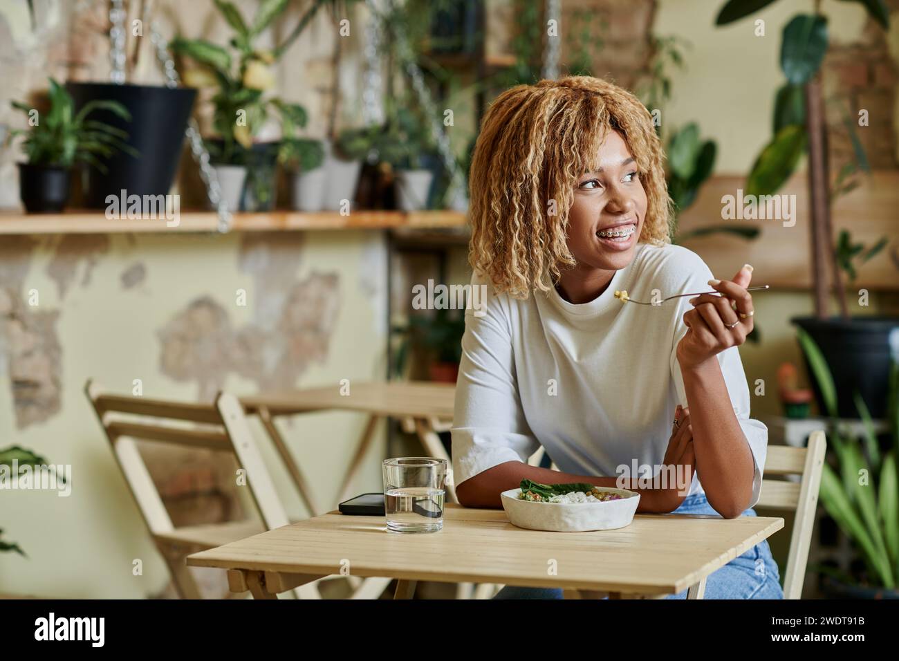 happy african american girl in braces eating fresh salad in bowl inside of plant-filled vegan ...