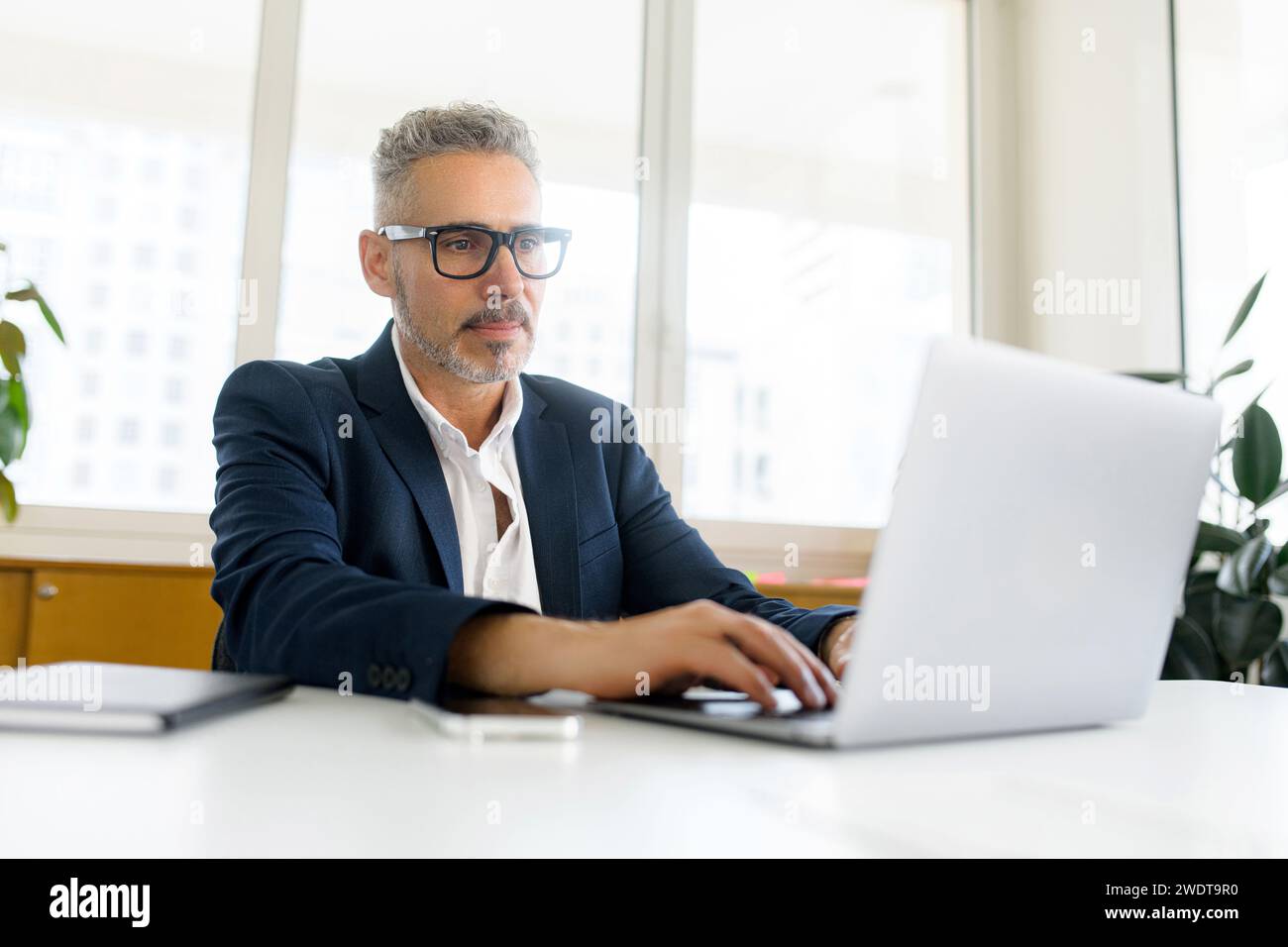 Focused mature senior businessman wearing eyeglasses using laptop in contemporary office space ...