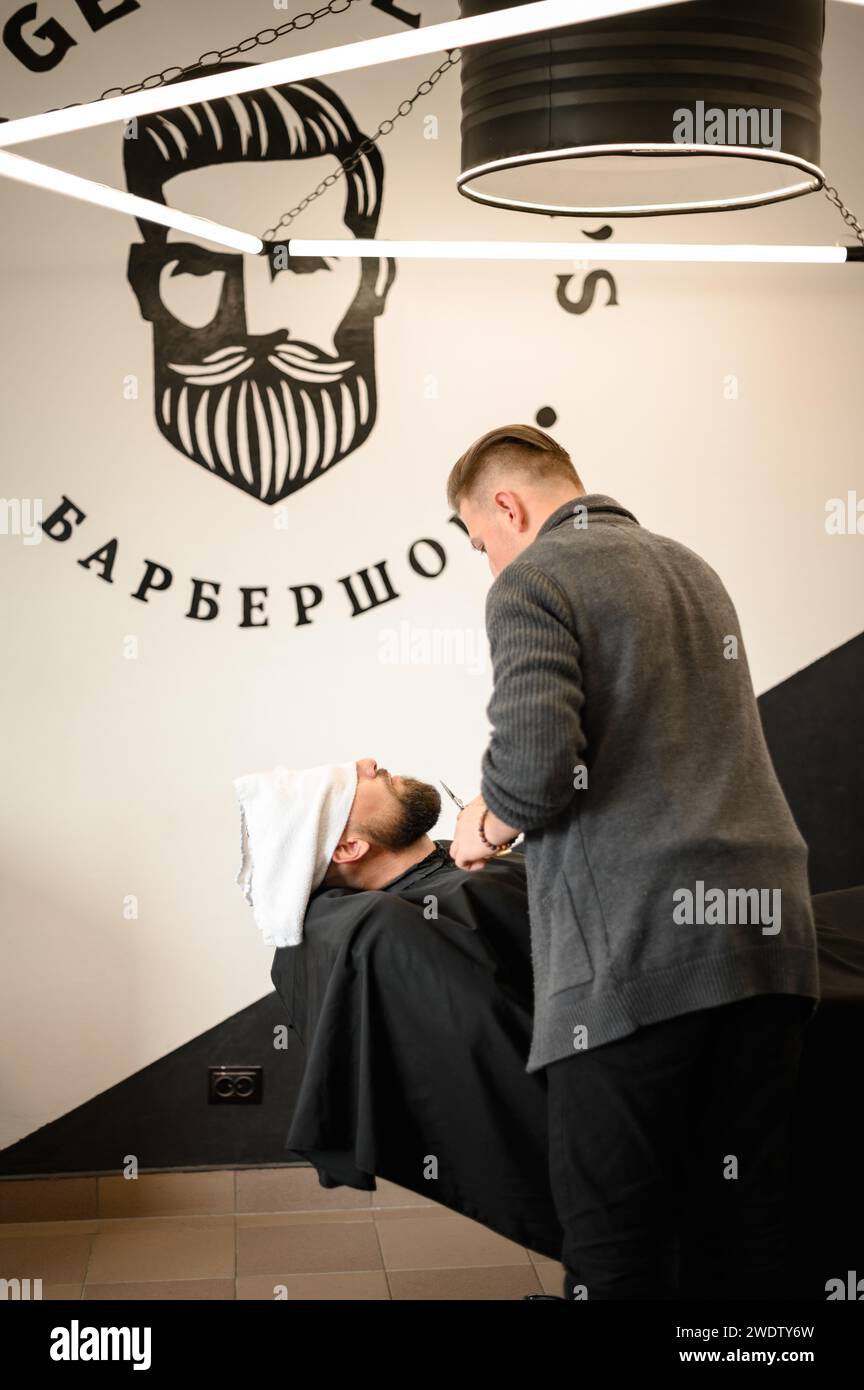 A barber stylist trims the beard of a Caucasian man, whose face is ...