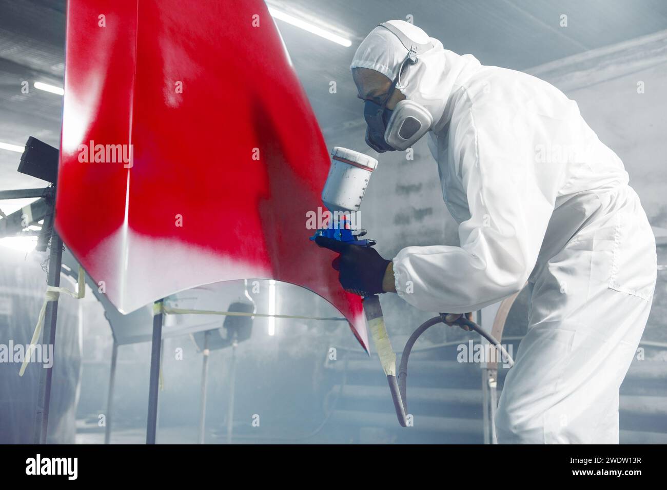 Worker paints car body part in workshop chamber using airbrush. Man ...
