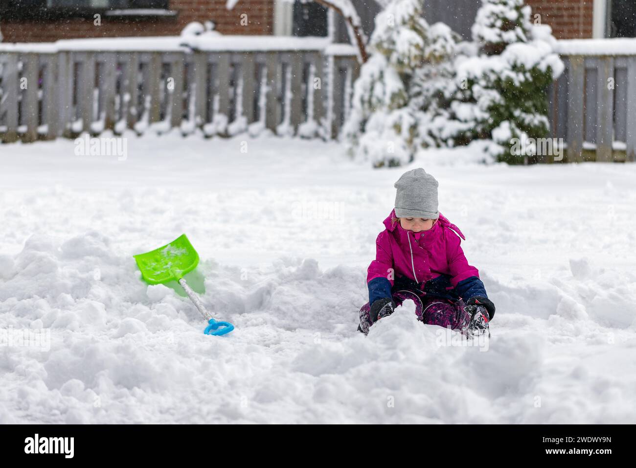 Little child playing with snow and shovel outdoors near house in winter ...