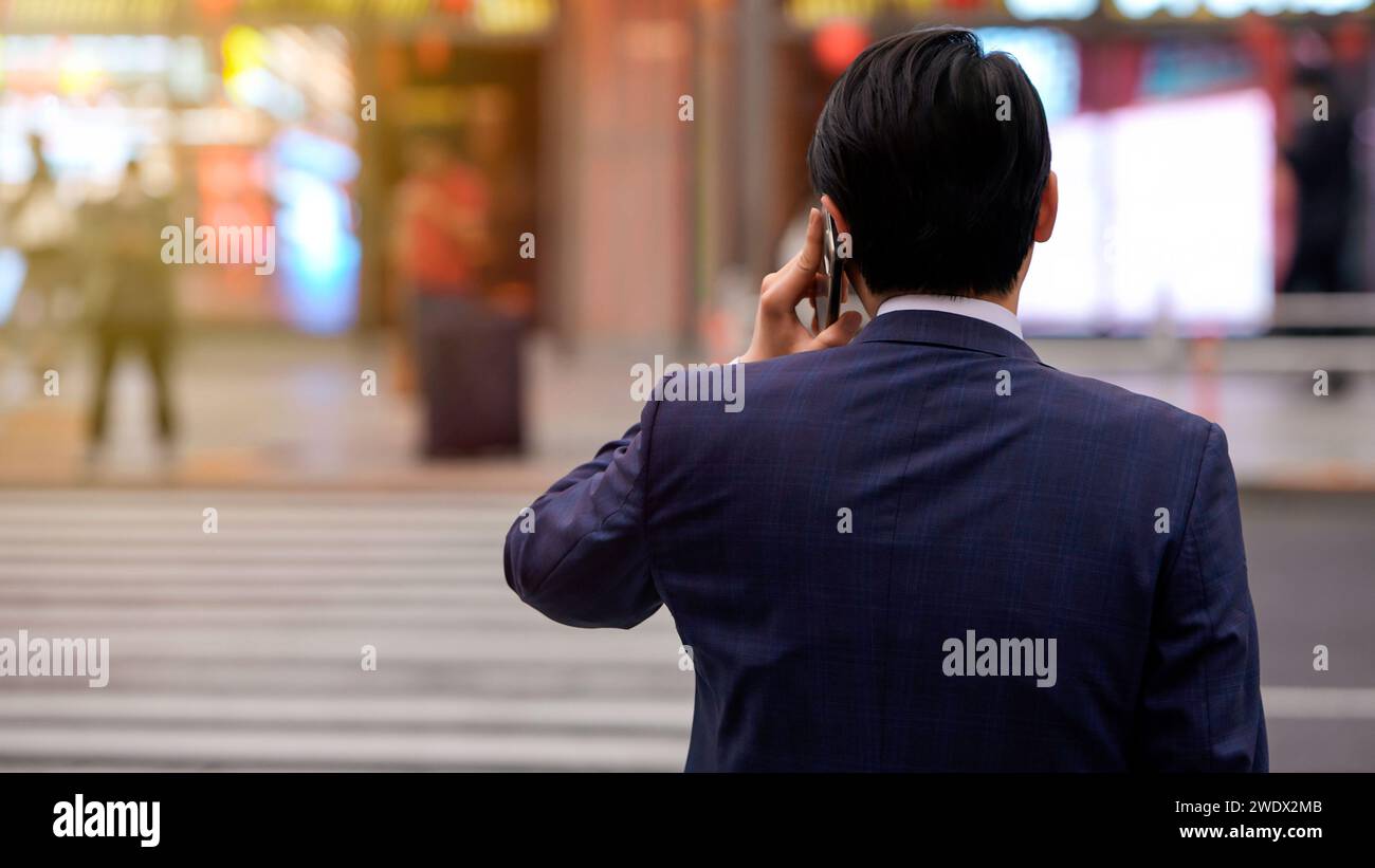 A Japanese businessman stands patiently in the crosswalk, using a smartphone in hand to make a ...