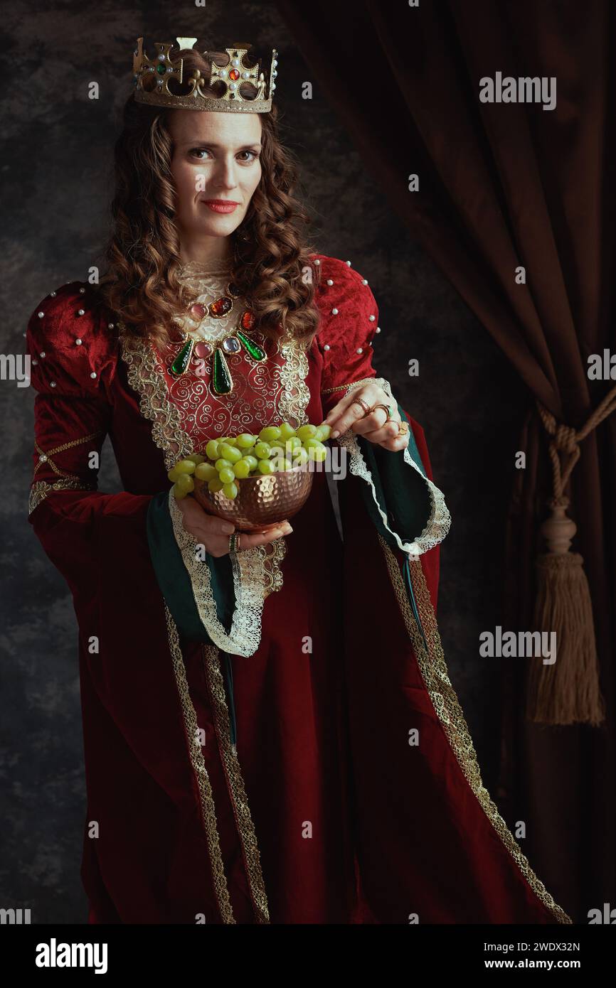 medieval queen in red dress with plate of grapes and crown on dark gray ...