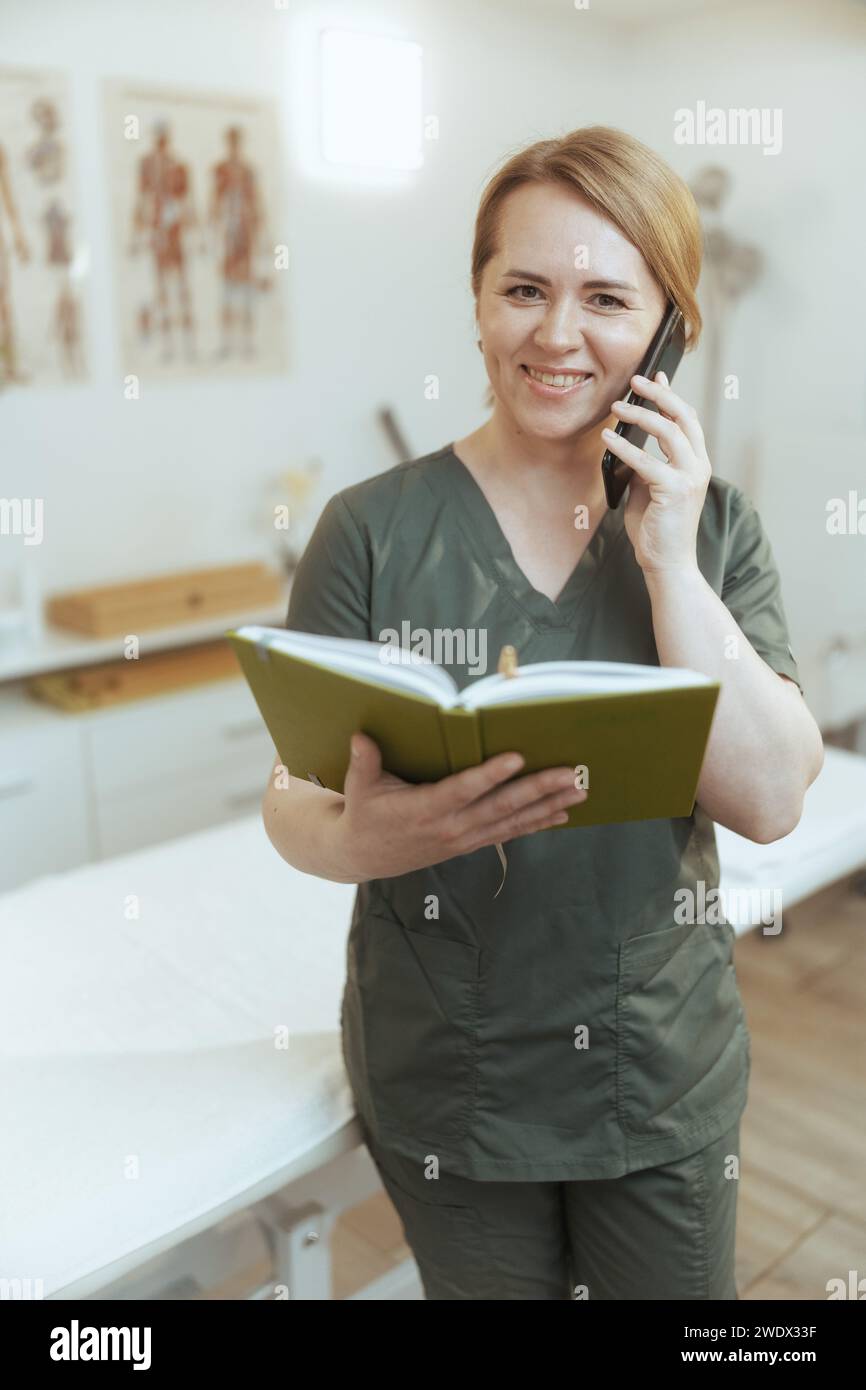 Healthcare time. happy female medical massage therapist in massage cabinet with notebook talking ...