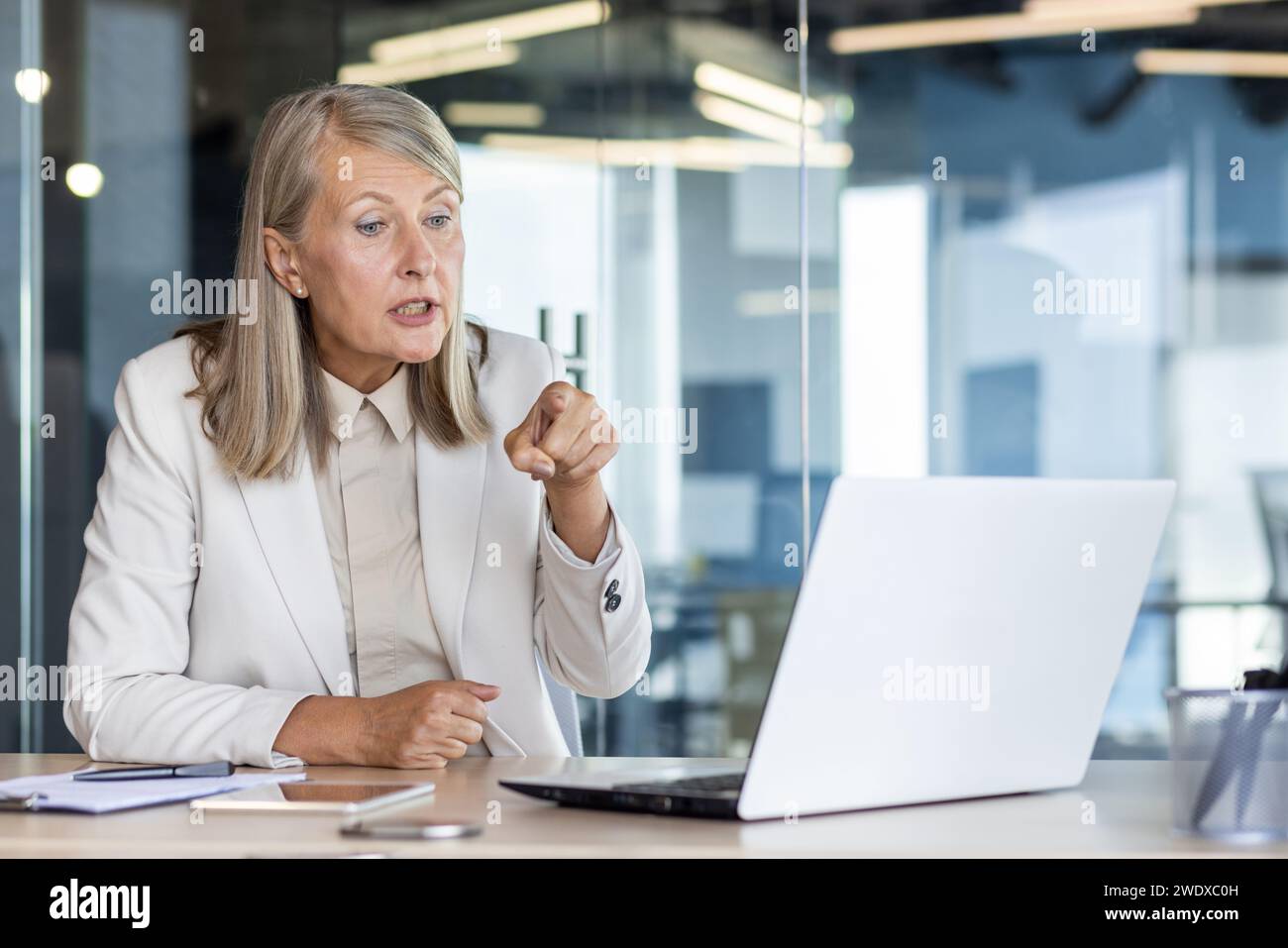 Angry senior business woman sitting in the office at the desk in a suit ...