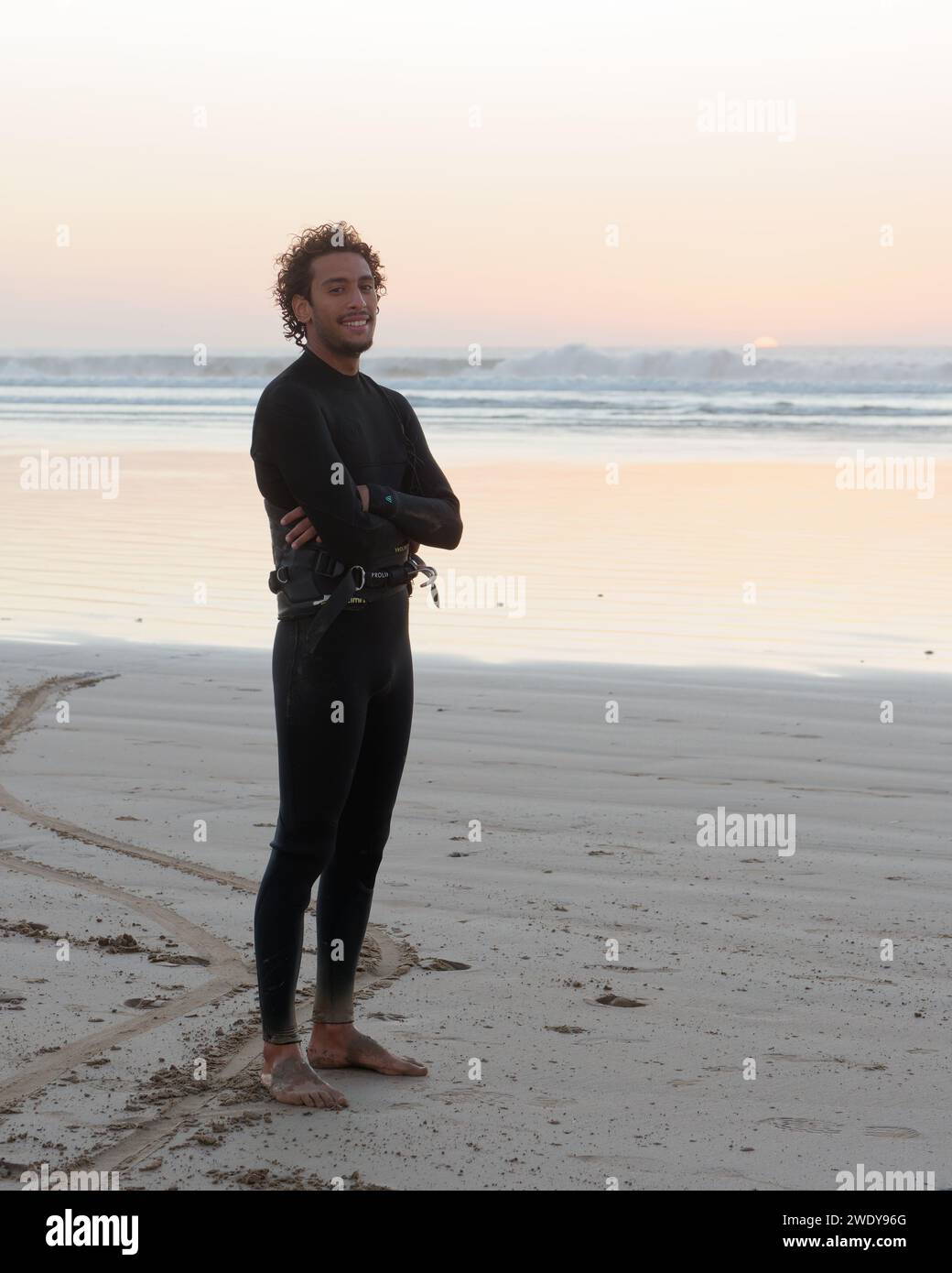 Handsome man in a wet suit with long hair poses for a photo on a sandy ...