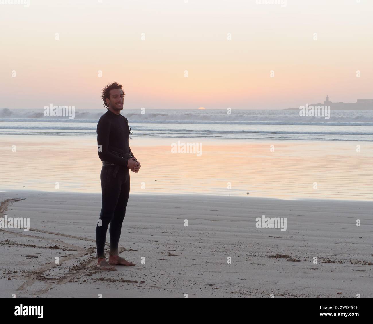 Handsome man in a wet suit with long hair poses for a photo on a sandy ...