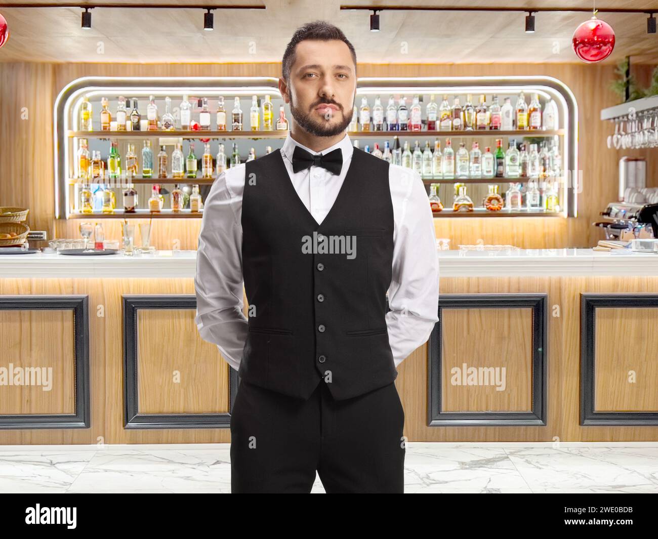 Waiter with a bow tie and vest standing in front of a bar Stock Photo - Alamy