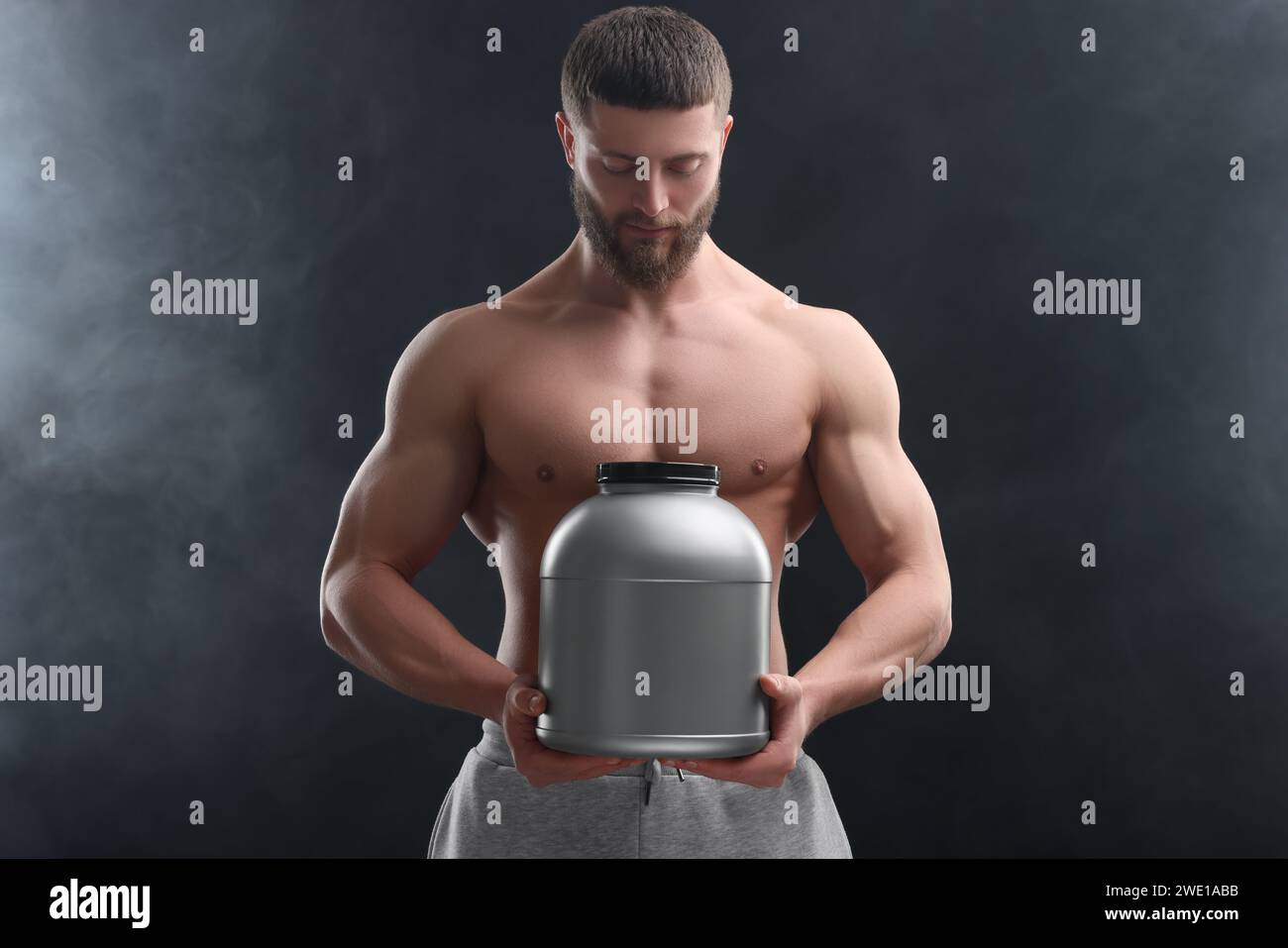 Young man with muscular body holding jar of protein powder on dark grey ...